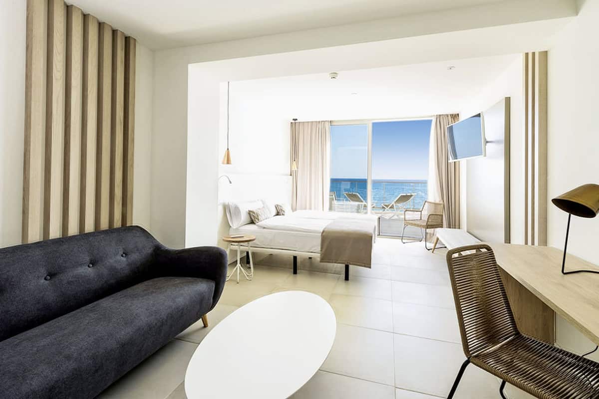 Quarto de hotel moderno com uma cama, sofá, escrivaninha e vista para o mar.