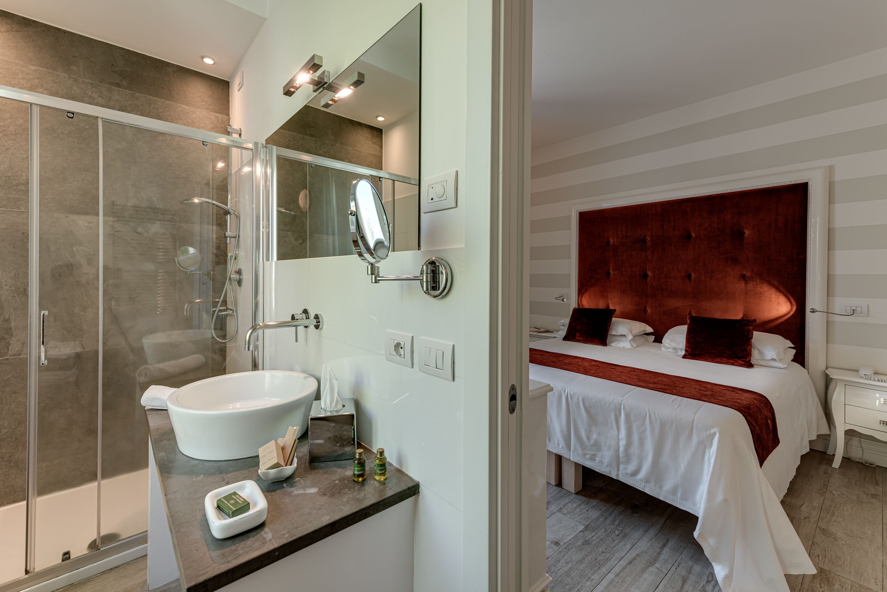 Casa de banho moderna com cabine de chuveiro em vidro, uma pia grande e um espelho. Junto, um quarto com uma cama grande, cabeceira vermelha e roupa de cama branca.