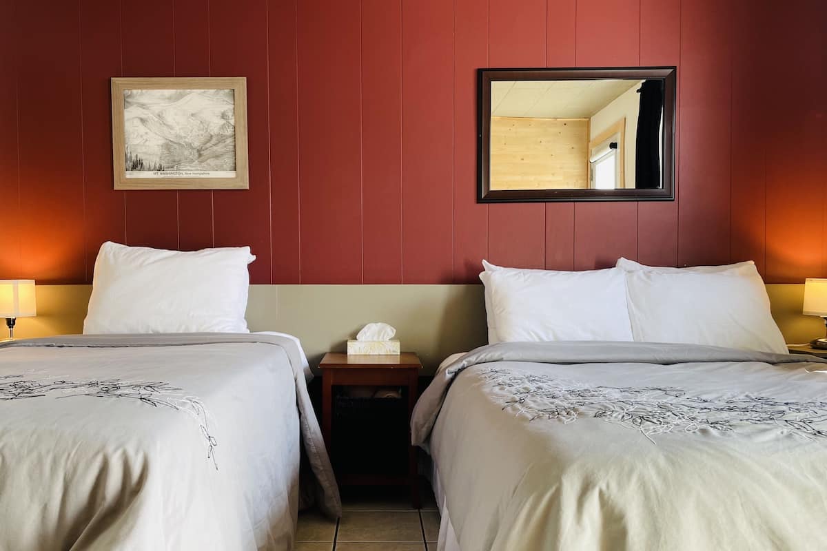 Quarto de hotel com duas camas, uma mesinha de cabeceira com abajur, um quadro na parede e um espelho sobre as camas.