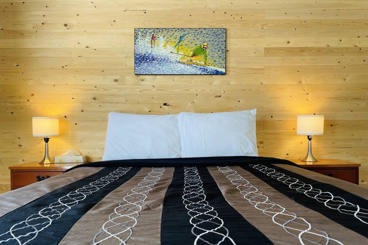 Uma cama bem arrumada com colcha estampada, duas luminárias de cabeceira, uma cabeceira de madeira e um quadro acima da cama.