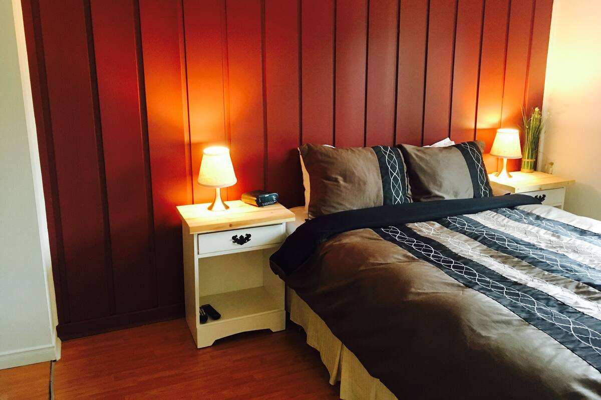 Quarto com cabeceira de madeira, cama com colcha estampada, duas mesas de cabeceira com abajures e um criado-mudo com controle remoto.
