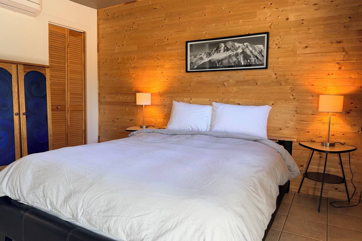Quarto com cabeceira de madeira, cama com roupa de cama branca, duas luminárias de mesa e um quadro na parede.