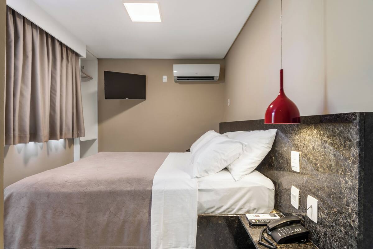 Quarto de hotel com cama, televisão na parede, aparelho de ar condicionado, telefone e luminária pendente vermelha.