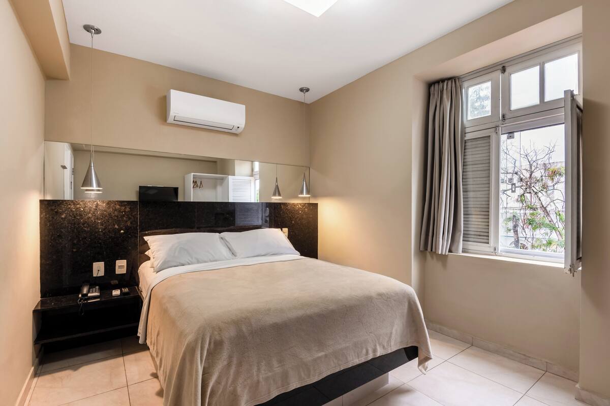 Quarto com cama, mesas de cabeceira, espelho e janela com cortinas.