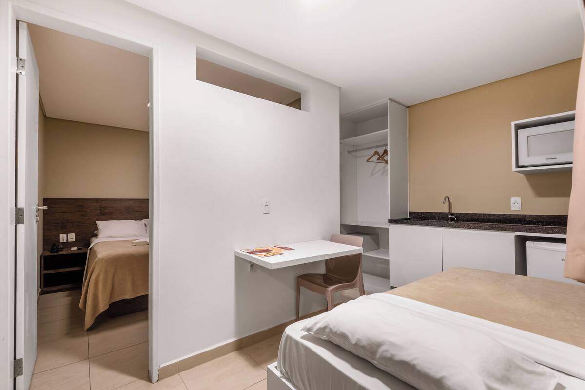 Quarto de hotel compacto com cama, pequena escrivaninha, cadeira, guarda-roupa e cozinha compacta.