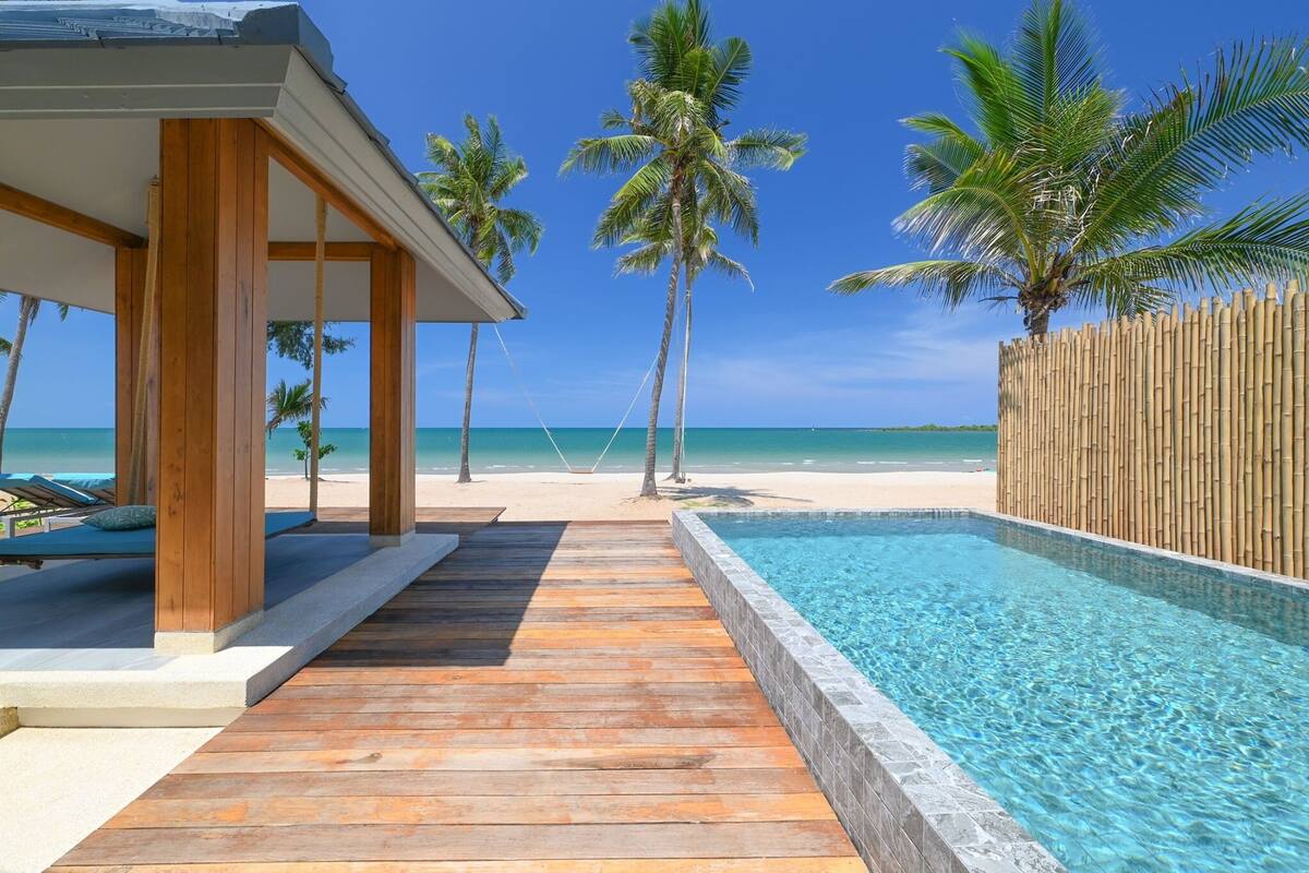 Um deck de madeira que leva a uma piscina, com cerca de bambu, palmeiras e vista para a praia.