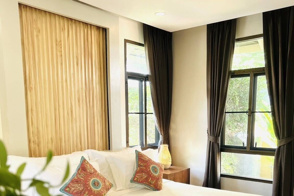 Quarto de hotel com uma cama grande, duas almofadas decorativas, uma mesa de cabeceira com abajur e uma janela com cortinas.