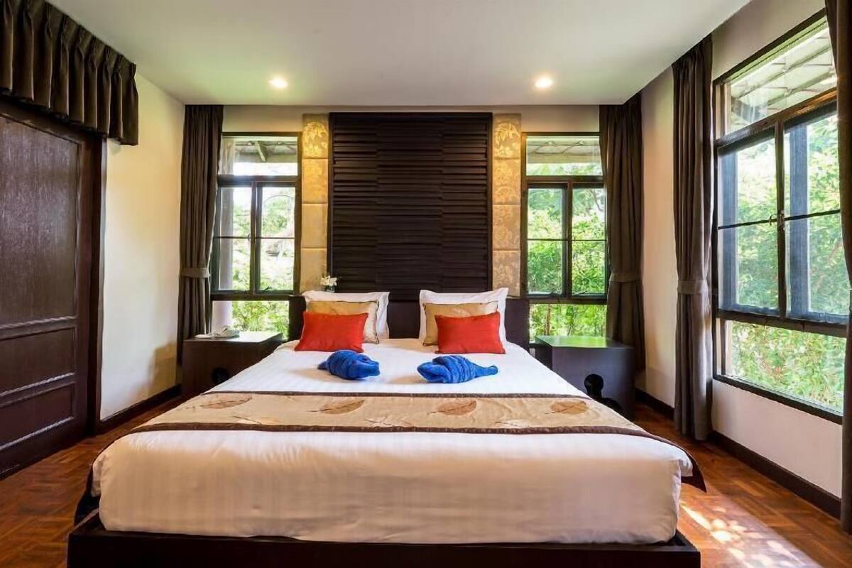 Uma cama grande com cabeceira de madeira, duas mesas de cabeceira e vista para o verde através de amplas janelas.
