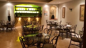 Uma área de refeições com mesas e cadeiras redondas, um espaço de bar com prateleiras cheias de garrafas e um vaso decorativo na parede.