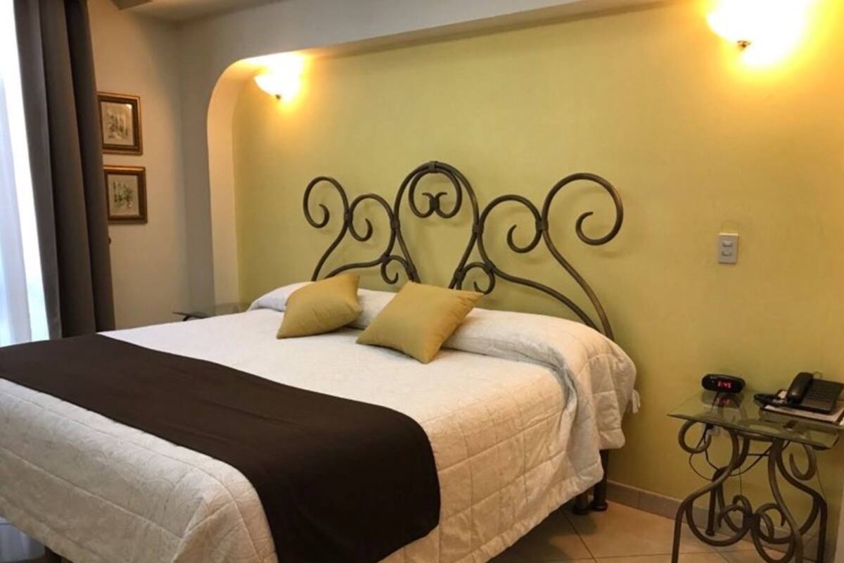 Uma cama bem arrumada com um cobertor preto, uma mesinha de cabeceira com um telefone e um relógio, e um aplique na parede.