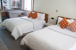Quarto de hotel com duas camas, cada uma com roupa de cama branca e almofadas laranja estampadas. Uma janela oferece vista para o exterior.