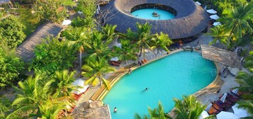 Vista aérea de um resort com uma grande piscina central, cercada por palmeiras e construções com telhados de palha.