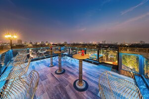 Um bar no terraço com cadeiras e mesas de metal, vista da cidade e guarda-corpo decorativo.