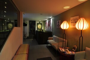 Uma sala de estar moderna com um sofá, uma mesa de centro com revistas, e duas luminárias fixadas na parede. Há relógios na parede e um espaço de bar ao fundo.