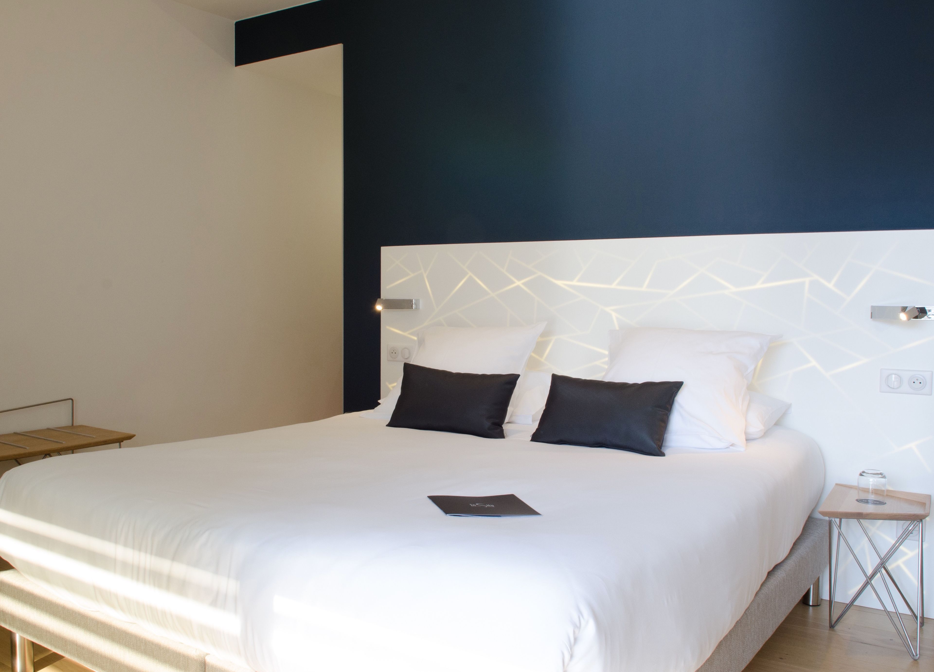 Quarto de hotel moderno com uma cama grande, uma mesa de cabeceira e um candeeiro fixo na parede.