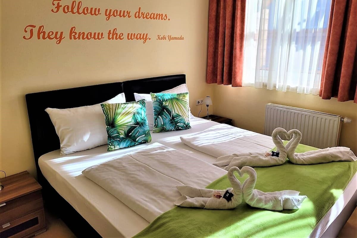 Uma cama bem arrumada com lençóis brancos, um cobertor verde e toalhas decorativas em formato de cisne. Uma frase motivacional está exposta na parede.