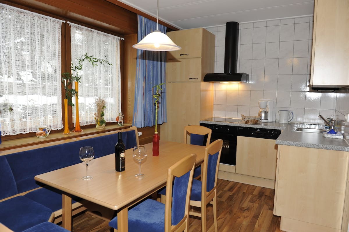 Uma cozinha compacta com armários de madeira, máquina de lavar louça embutida e um espaço de jantar com cadeiras estofadas em azul e mesa de madeira.