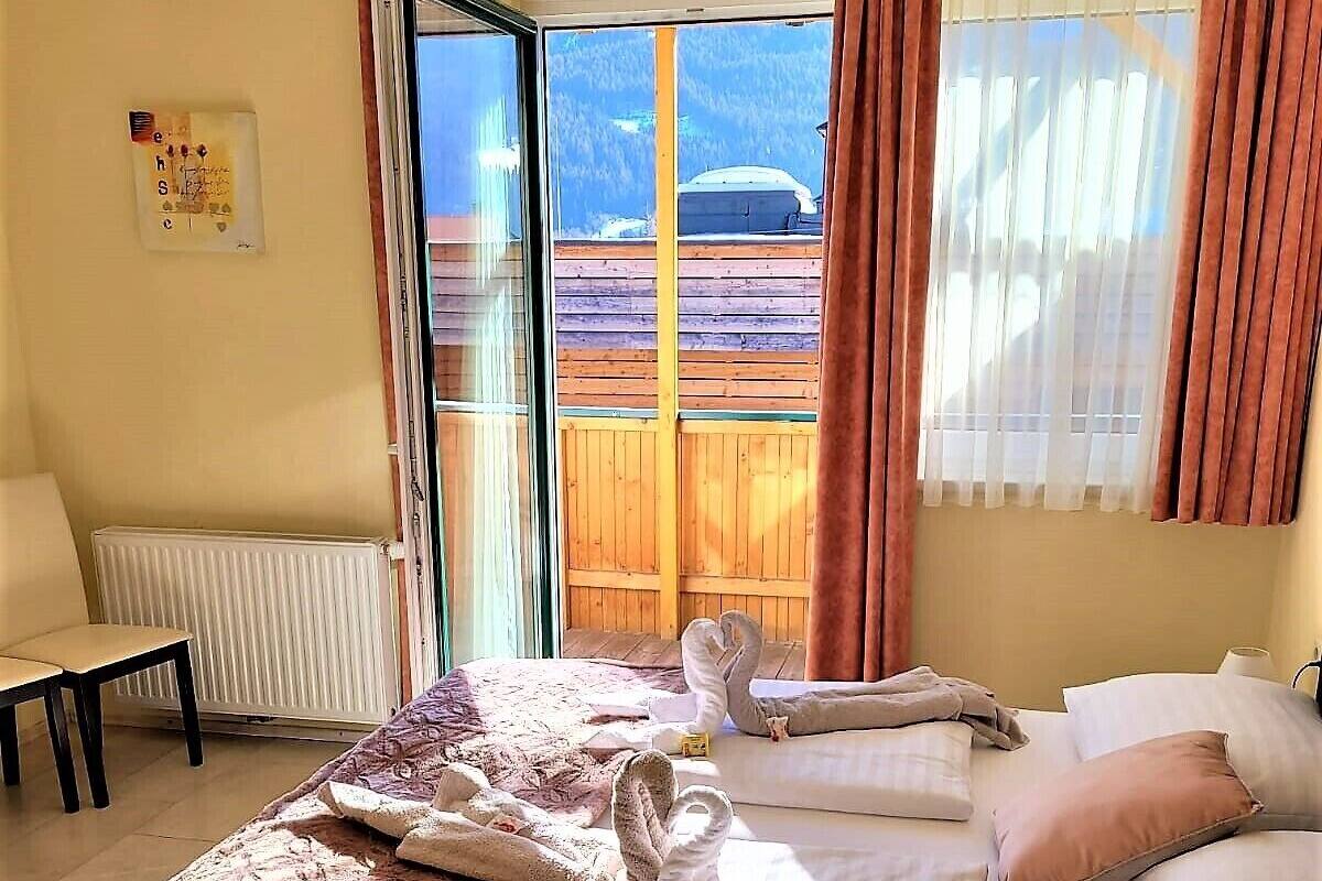 Quarto com cama, escrivaninha e vista para um deck de madeira e montanhas.