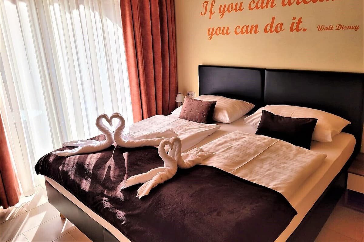 Cama arrumada com decoração de toalha em formato de coração, uma frase motivacional na parede e uma janela com cortinas.