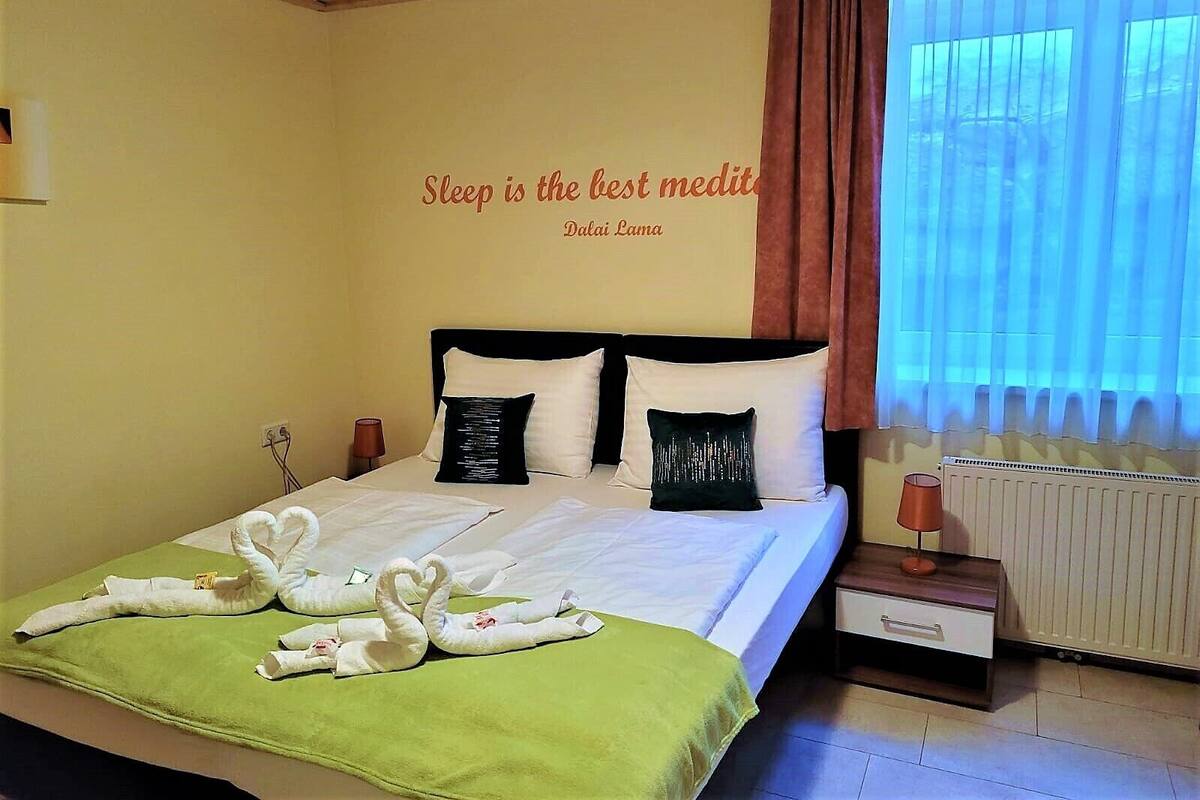 Uma cama bem arrumada com almofadas brancas e pretas, um cobertor verde e duas toalhas em formato de cisne. Uma frase na parede diz: 'O sono é a melhor meditação'.