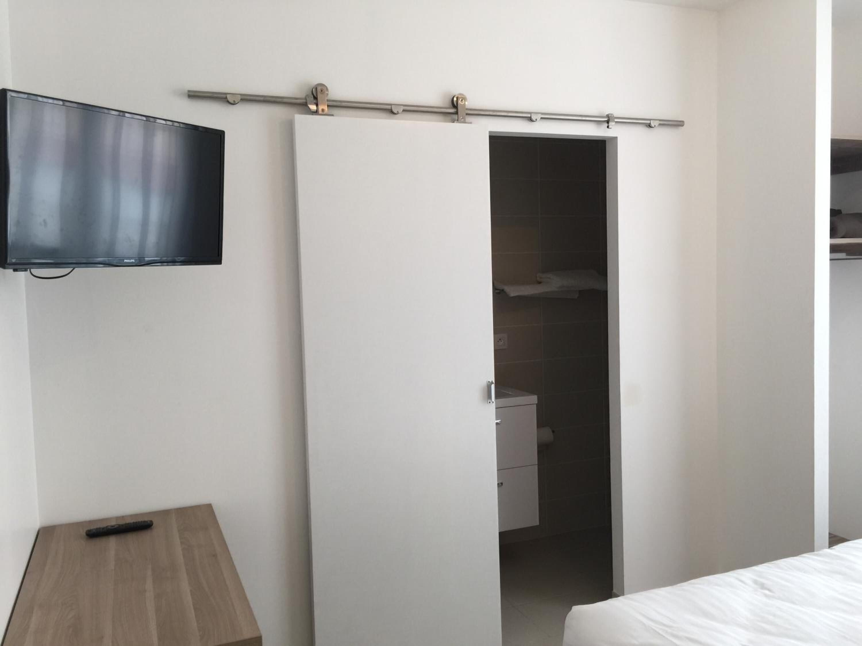 Quarto de hotel com uma cama, uma televisão fixada na parede, uma mesa de cabeceira de madeira e uma porta que dá para o banheiro.