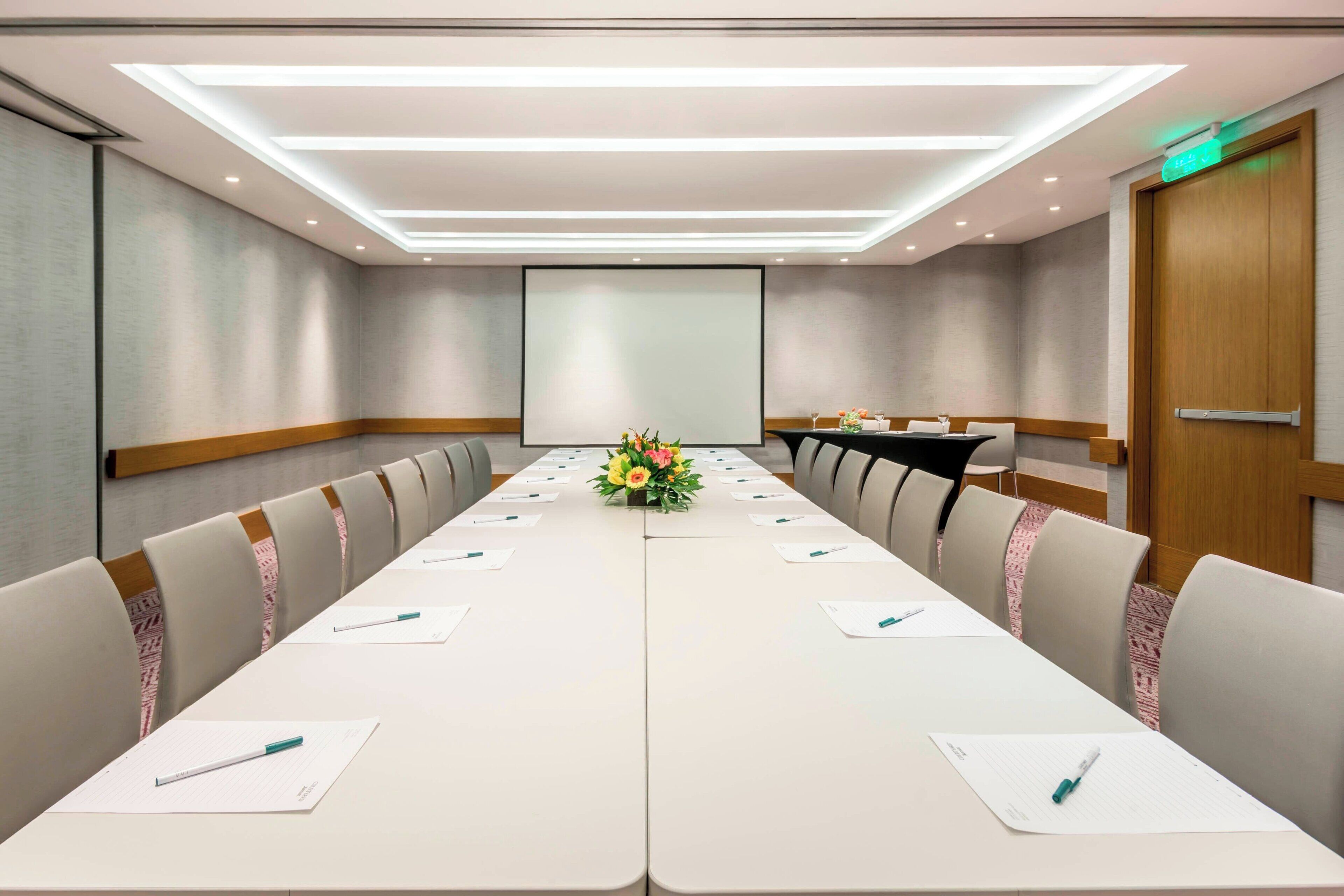 Uma sala de conferência com uma mesa comprida, cadeiras, tela de projetor e um arranjo floral.