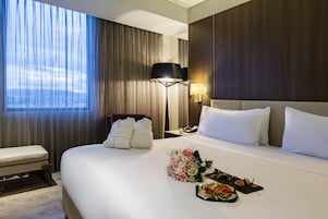 Quarto de hotel com uma cama grande, uma mesa de cabeceira com abajur, um banco, um buquê de flores e uma bandeja com comida.