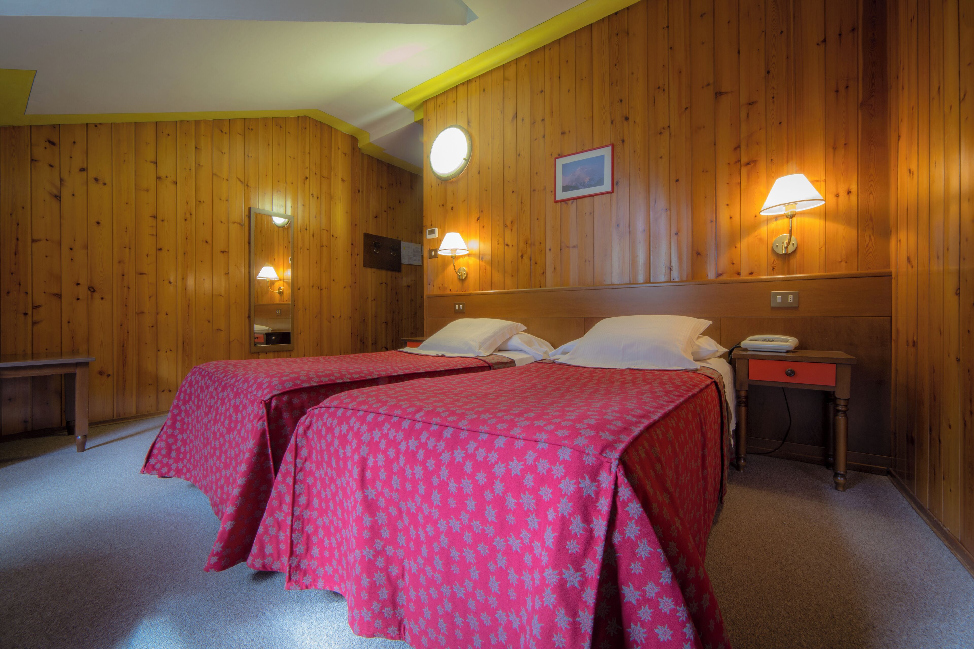 Quarto com paredes de painel de madeira, duas camas com roupa de cama floral rosa, luminárias de cabeceira, mesa de cabeceira pequena e um espelho.
