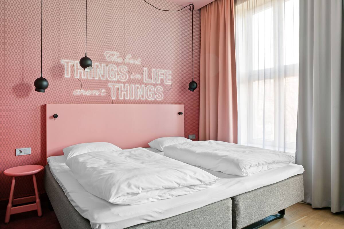 Quarto com parede rosa, cama com roupa de cama branca, um banquinho rosa pequeno e luminária no teto.