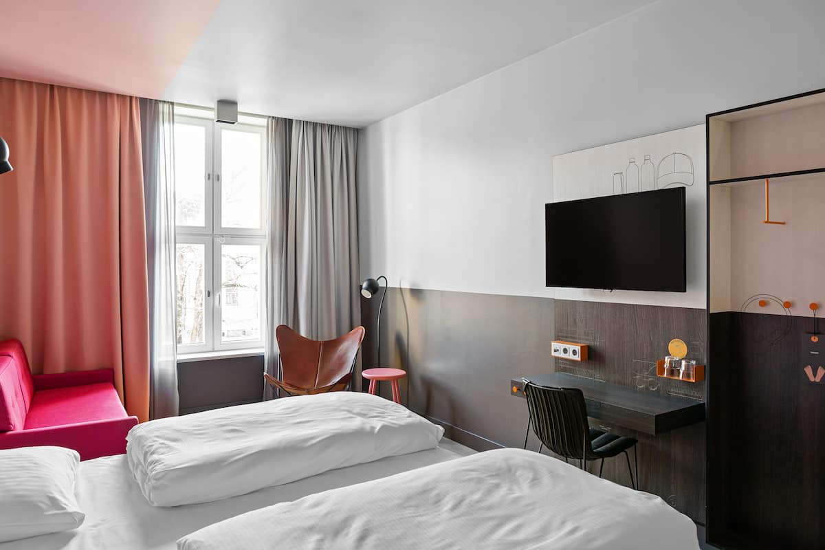 Quarto de hotel com duas camas, uma escrivaninha, uma cadeira, uma televisão e uma janela com cortinas.
