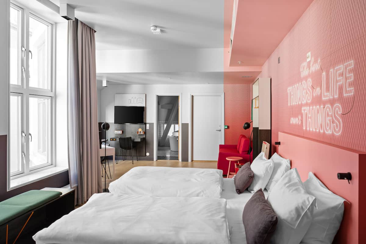 Quarto de hotel moderno com uma cama grande, uma escrivaninha e uma parede de destaque vermelha.