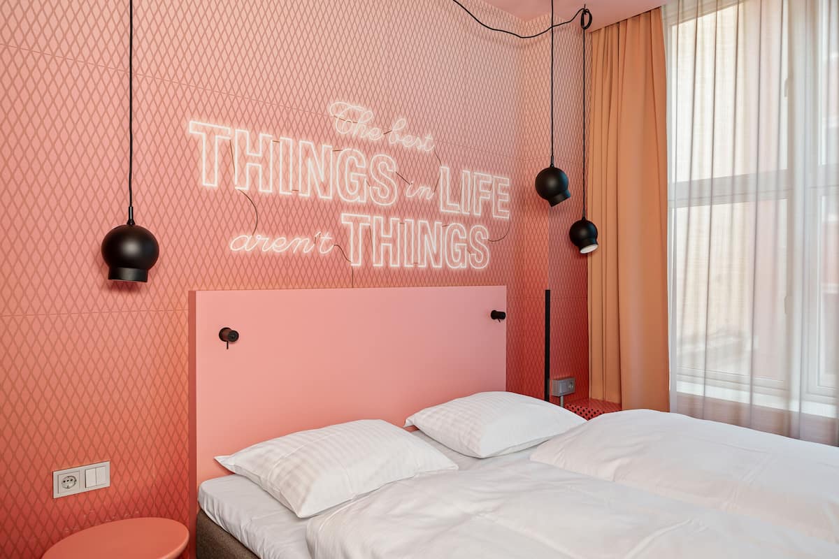 Quarto de hotel em tons de rosa, com uma cama, duas mesas de cabeceira e um letreiro de neon na parede.