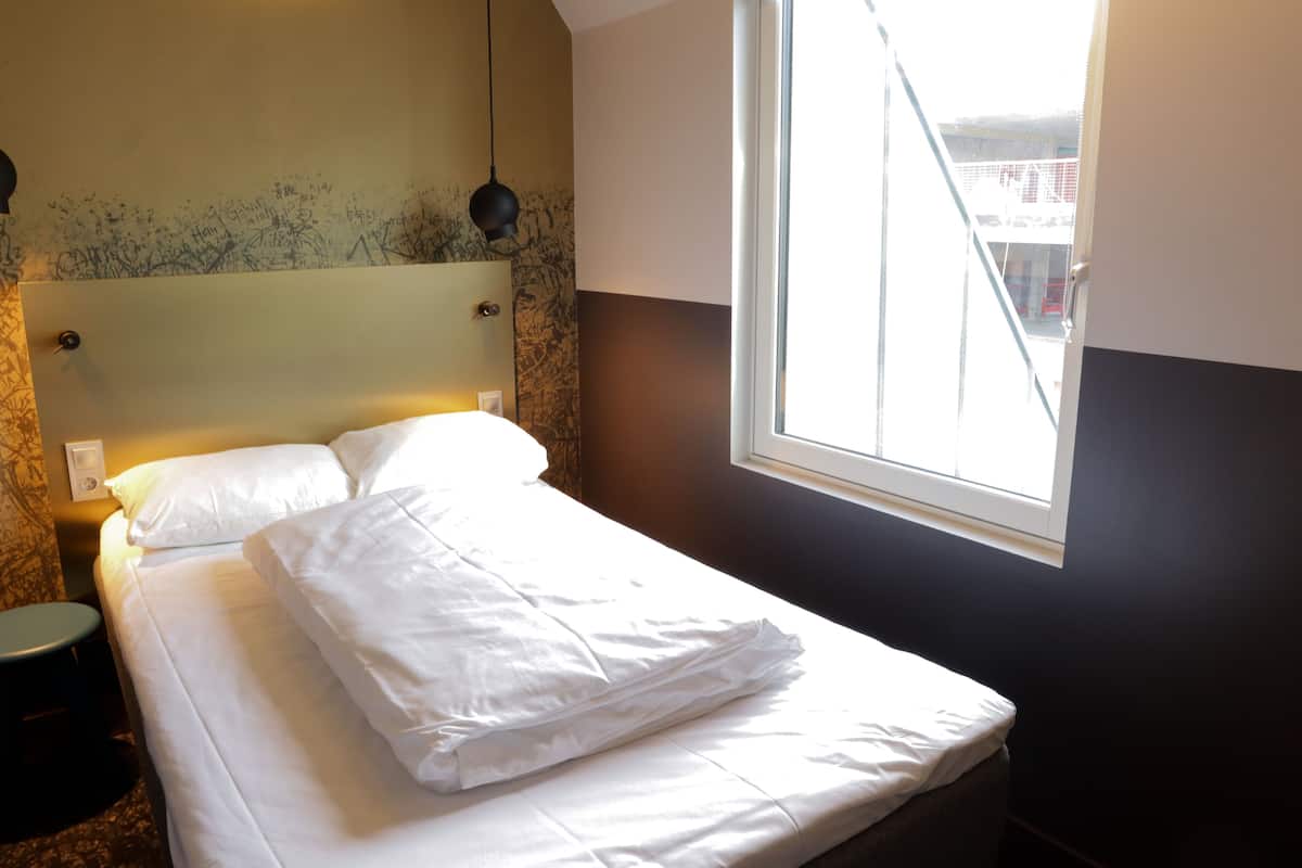 Cama bem arrumada em quarto com janela ampla, parede com grafite e luminária suspensa.
