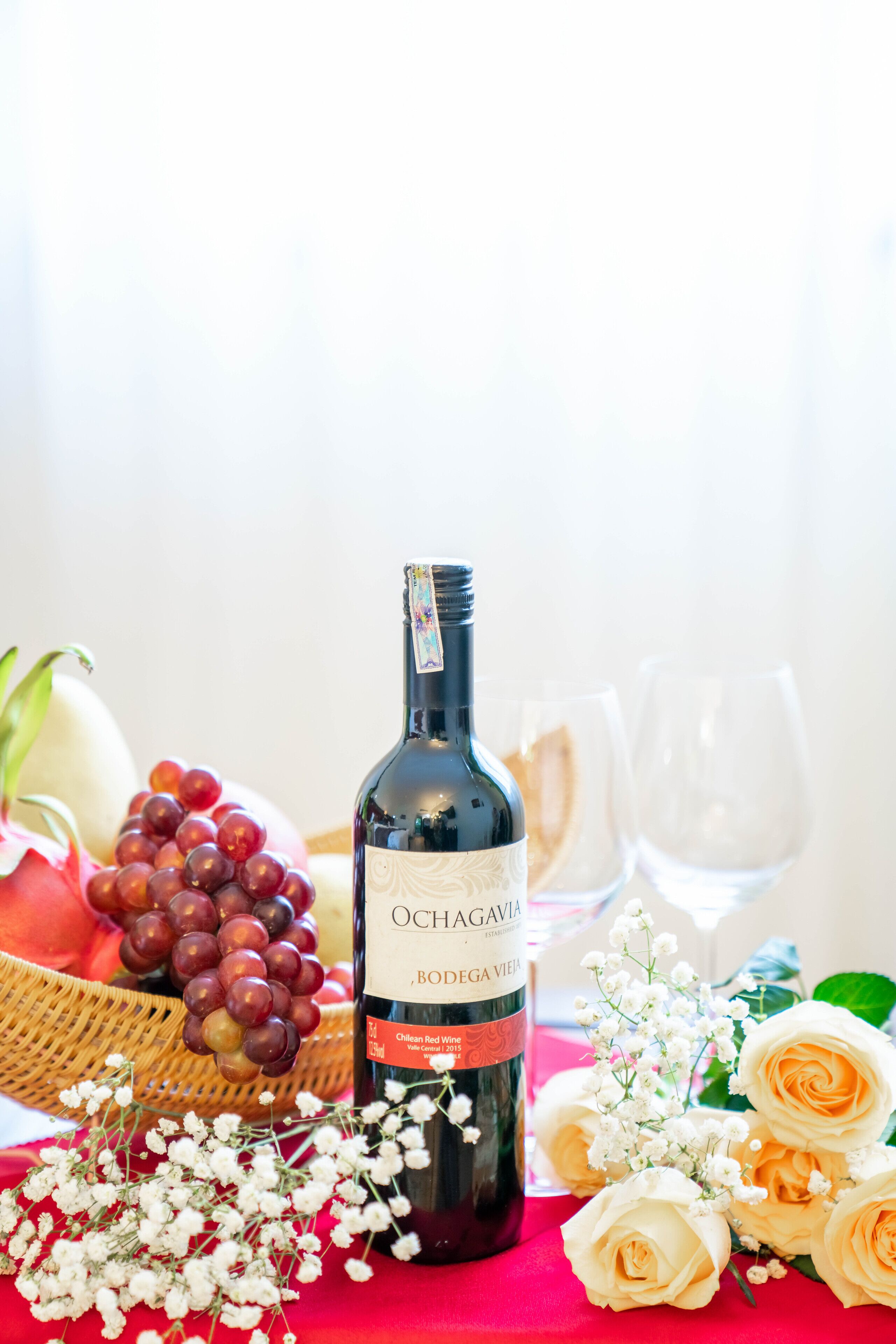 Uma garrafa de vinho Ochagavia Bodega Galicia, uma cesta de uvas, uma melancia e algumas flores sobre uma mesa.