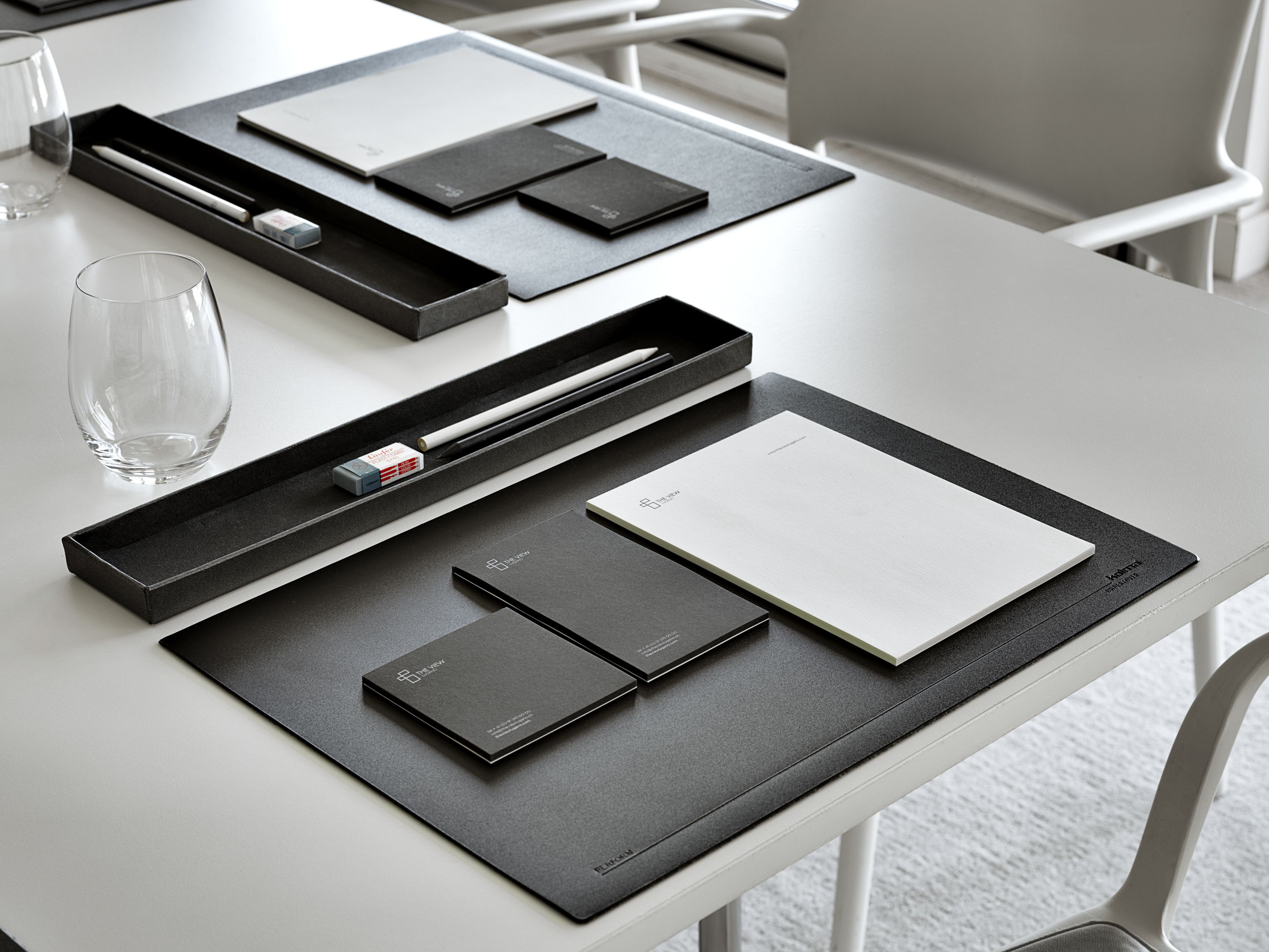 Uma secretária organizada com mesa branca, bandejas pretas, um copo, uma caneta e um bloco de notas.