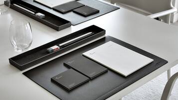 Uma secretária organizada com mesa branca, bandejas pretas, um copo, uma caneta e um bloco de notas.