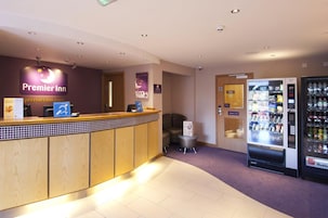 Uma área de recepção de hotel com um balcão de madeira, a placa da Premier Inn, uma área de estar e máquinas de venda automática.