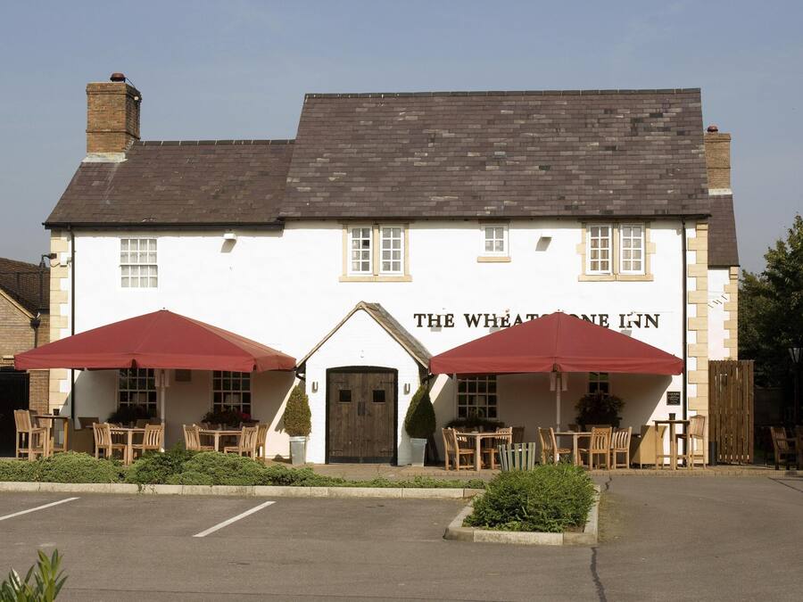 Um edifício branco com uma placa que diz 'The Wheatone Inn'. Há dois guarda-sóis vermelhos a oferecer sombra para o espaço de refeições ao ar livre.