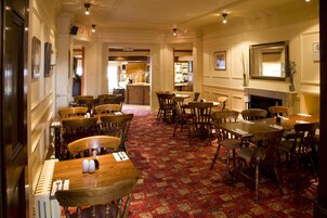 Uma área de refeições de hotel com várias mesas e cadeiras de madeira, um tapete estampado e um bar ao fundo.