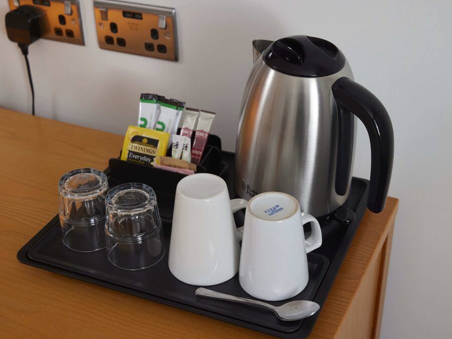 Uma bandeja com uma chaleira de aço inoxidável, duas canecas brancas, duas taças pequenas, uma colher e uma embalagem de saquinhos de chá Twinings.