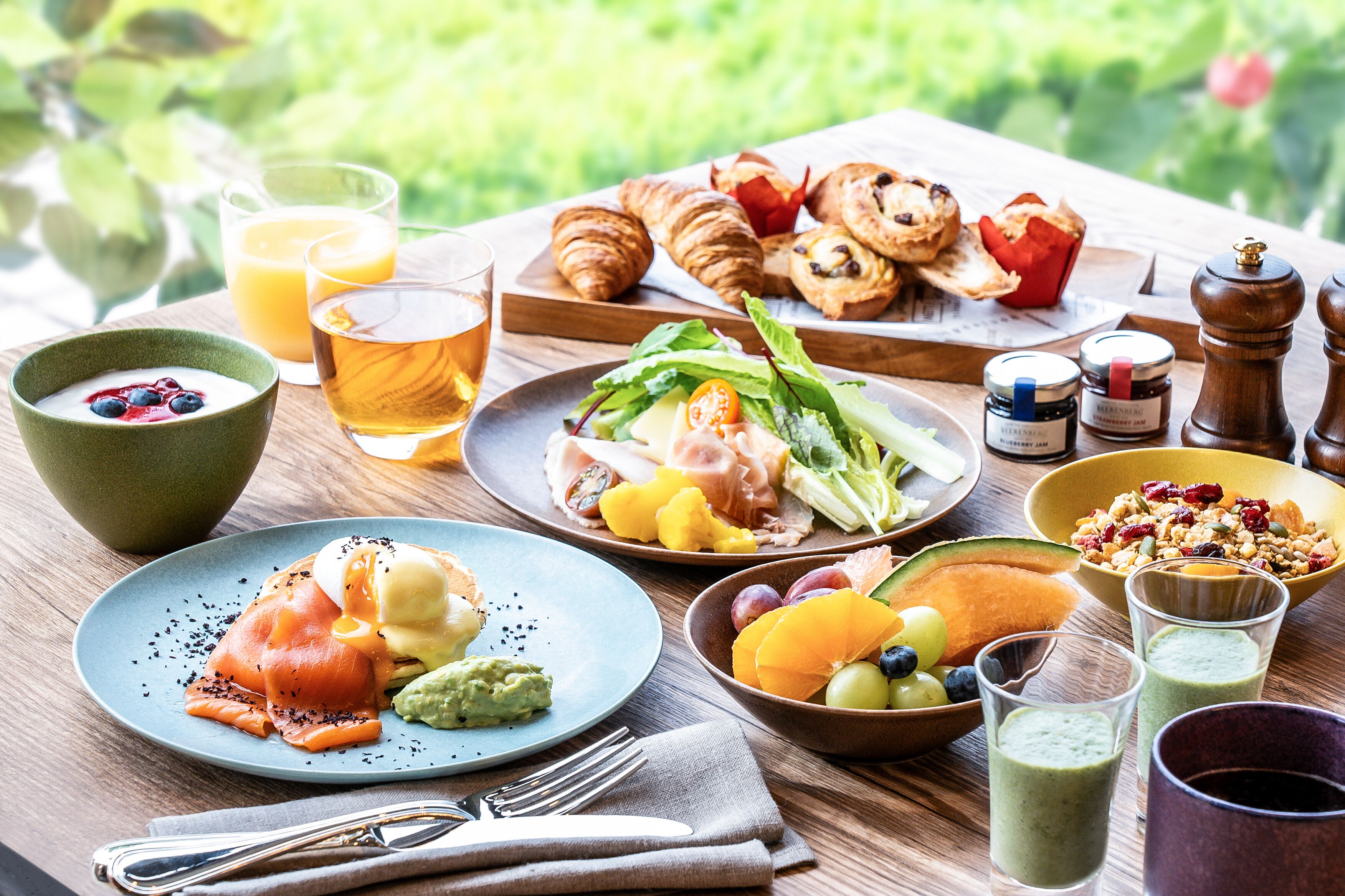 Disposição de café da manhã com croissants, salmão defumado, abacate e diversas frutas e sucos sobre uma mesa de madeira.