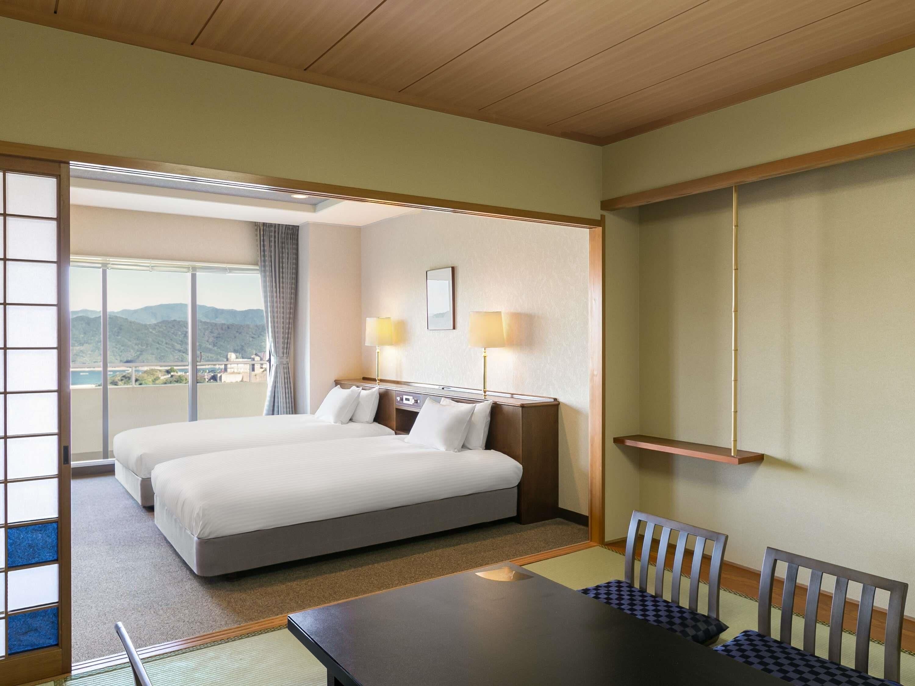 Quarto de hotel com uma cama, duas luminárias de cabeceira, uma mesa com cadeiras e vista para o exterior.