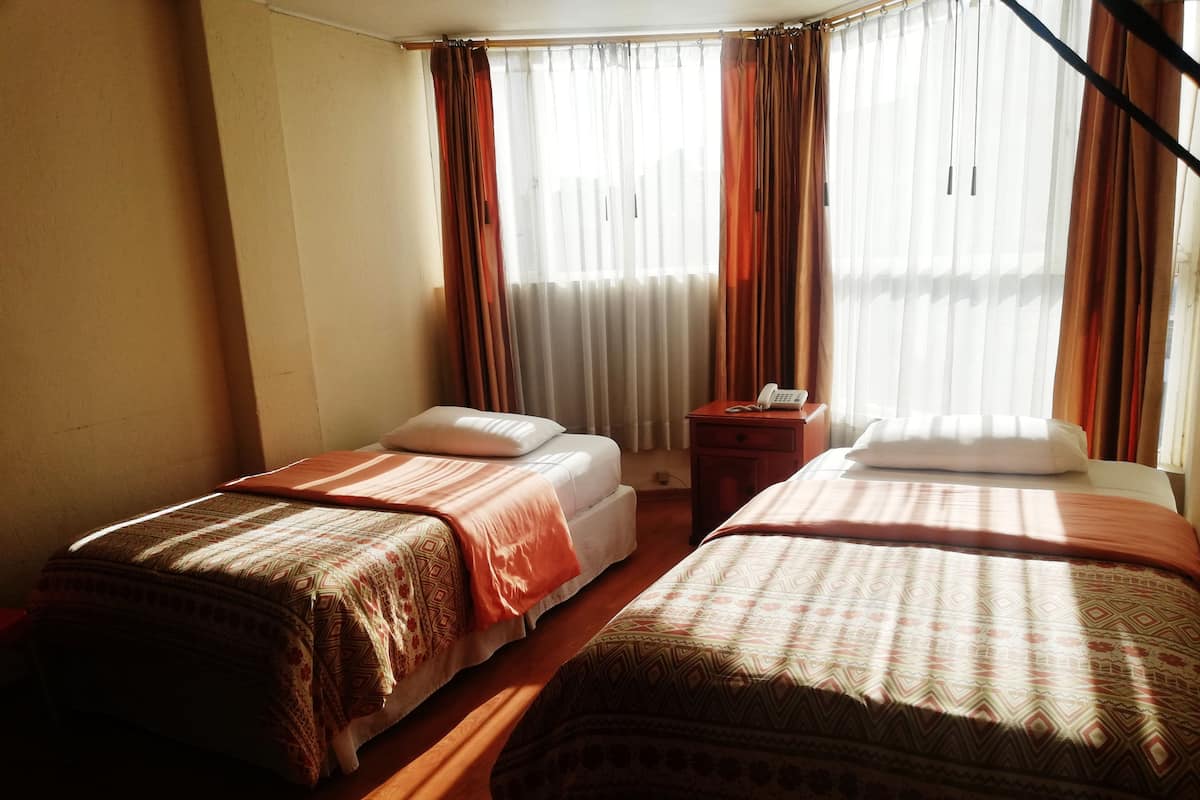 Quarto de hotel com duas camas, uma mesa de cabeceira com telefone e uma janela com cortinas.
