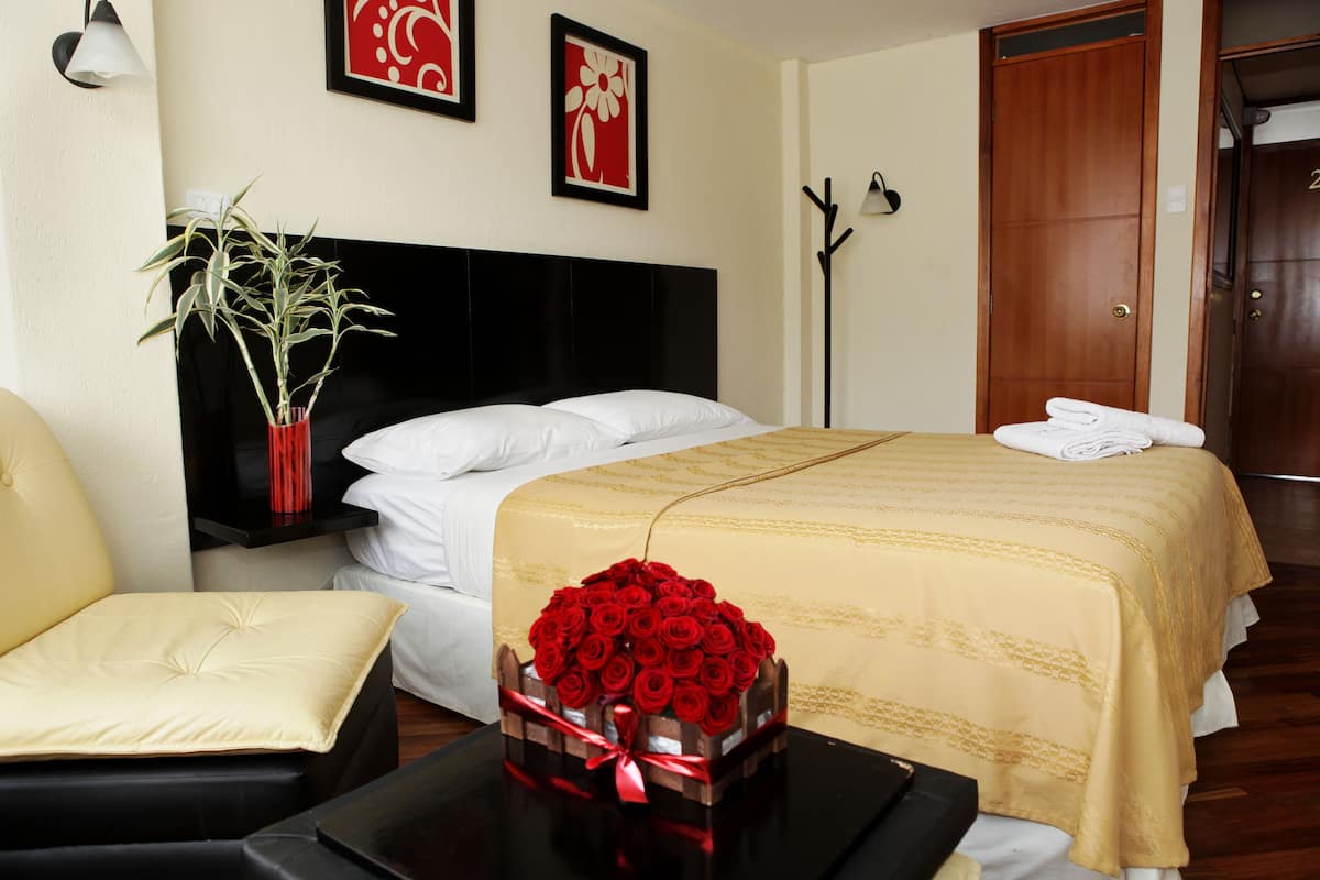 Quarto de hotel com cama, mesa de cabeceira com um vaso de rosas vermelhas, uma cadeira e um criado-mudo.