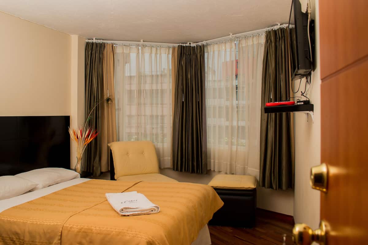 Quarto de hotel com cama, cadeira, televisão e janela com cortinas.
