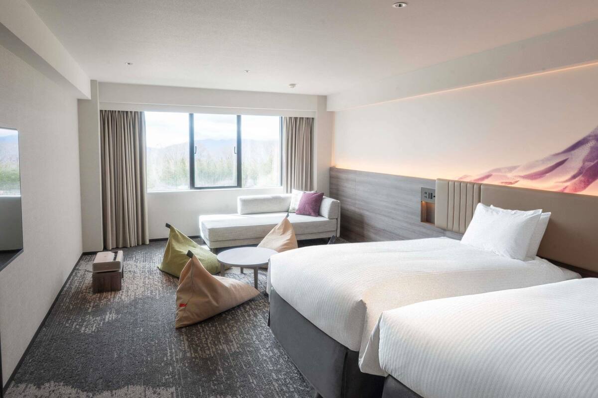 Quarto de hotel com cama, duas áreas de estar, janela com cortinas e luminária fixada na parede.
