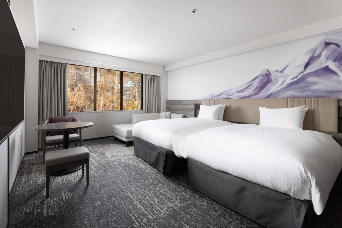 Quarto de hotel com uma cama grande, uma escrivaninha, uma cadeira e vista para árvores de outono pela janela.