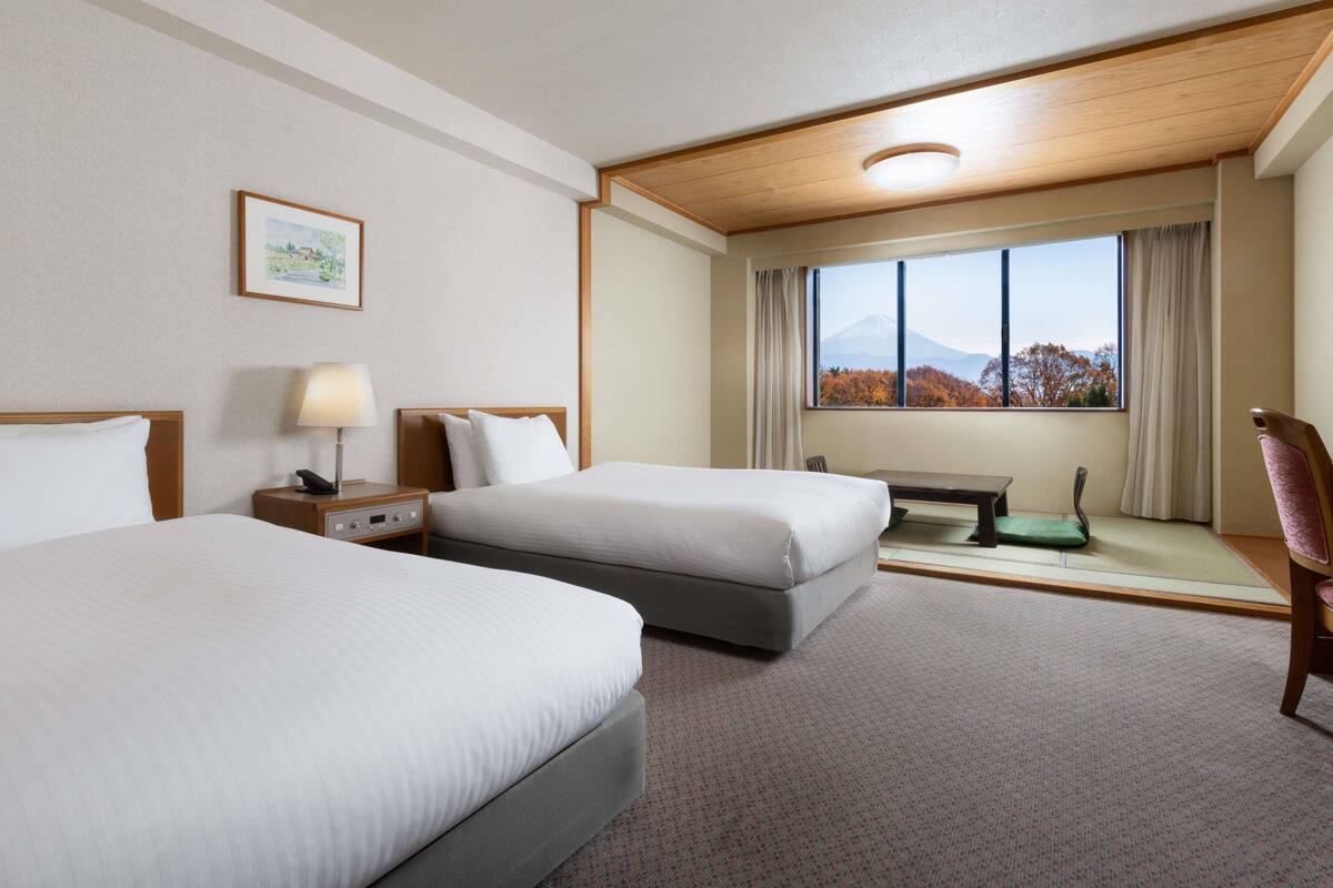 Quarto de hotel com duas camas, uma escrivaninha, uma cadeira, um abajur, um quadro na parede e uma janela com vista para uma montanha.