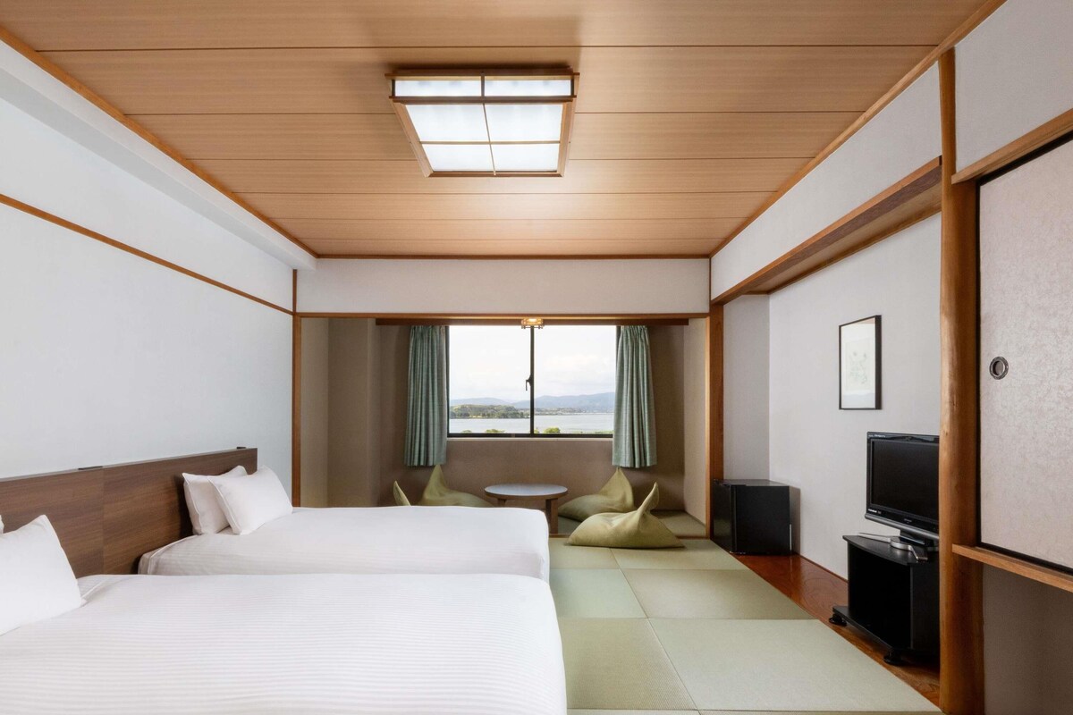 Quarto de hotel com duas camas, um espaço de estar pequeno, televisão e vista para o exterior.