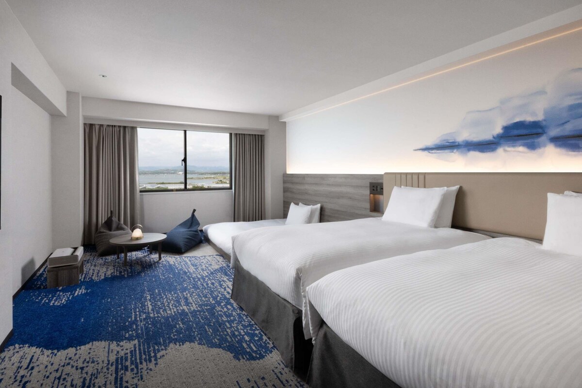 Quarto de hotel com duas camas, uma mesinha com uma cadeira, vista para o exterior e um quadro na parede.